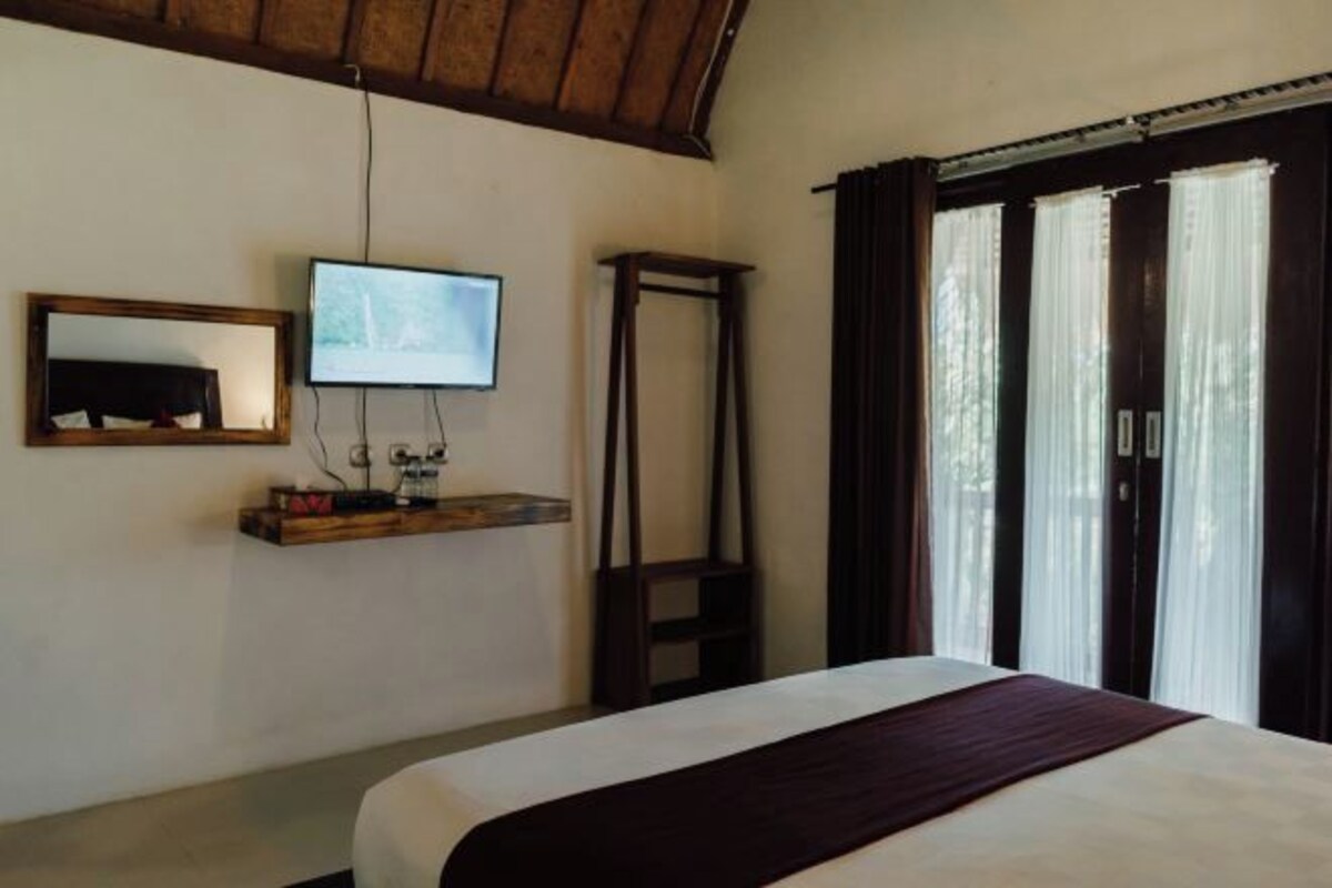 Quarto com cama, televisão montada na parede, prateleira de madeira e janela com cortinas.