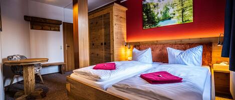 Quarto de hotel com cama de madeira, uma pequena mesa e um quadro de uma floresta na parede.