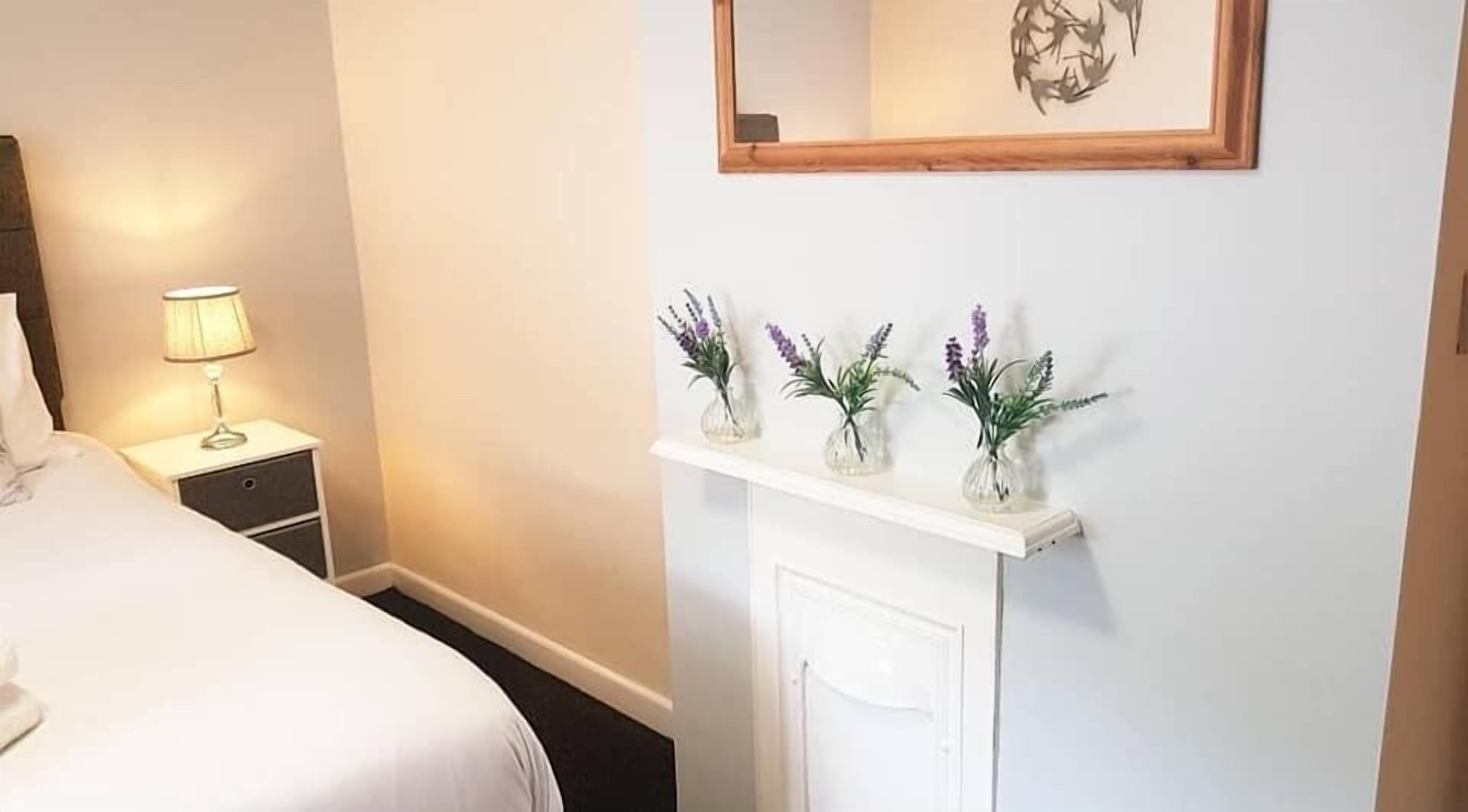 Quarto de hotel com cama, mesa de cabeceira com abajur e lareira com três vasos de lavanda.