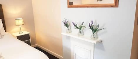 Quarto de hotel com cama, mesa de cabeceira com abajur e lareira com três vasos de lavanda.