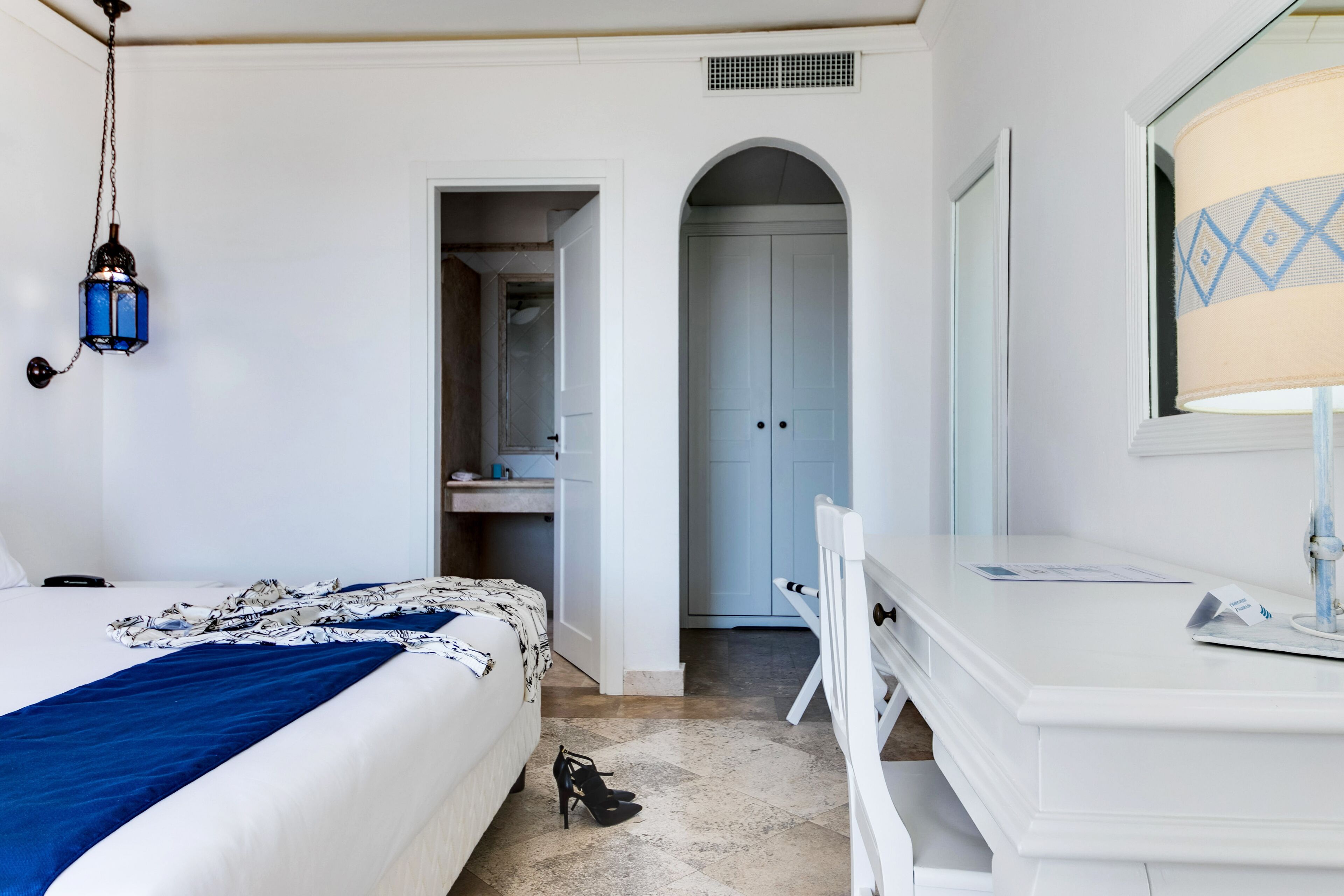Quarto de hotel com uma cama, uma escrivaninha, uma cadeira, um espelho e uma porta que dá para um banheiro.