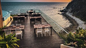 Um deck de madeira com mesas e cadeiras oferece vista para a área costeira, com piscina e uma linha costeira rochosa.