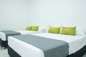 Quarto de hotel com duas camas de solteiro, cada uma com colcha cinza e travesseiro verde com a etiqueta "Ayenda Hotels".