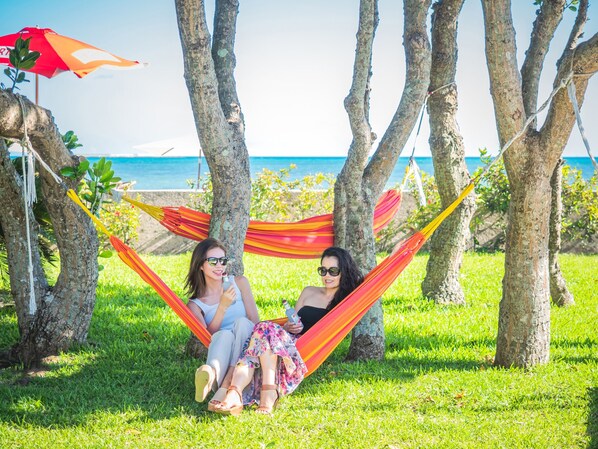 Duas pessoas relaxando em uma rede entre árvores em uma área gramada, com vista para o oceano.