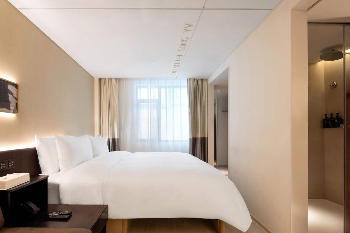 Quarto de hotel com uma cama grande, uma mesa de cabeceira com controle remoto, um criado-mudo, uma janela com cortinas e uma área de chuveiro.