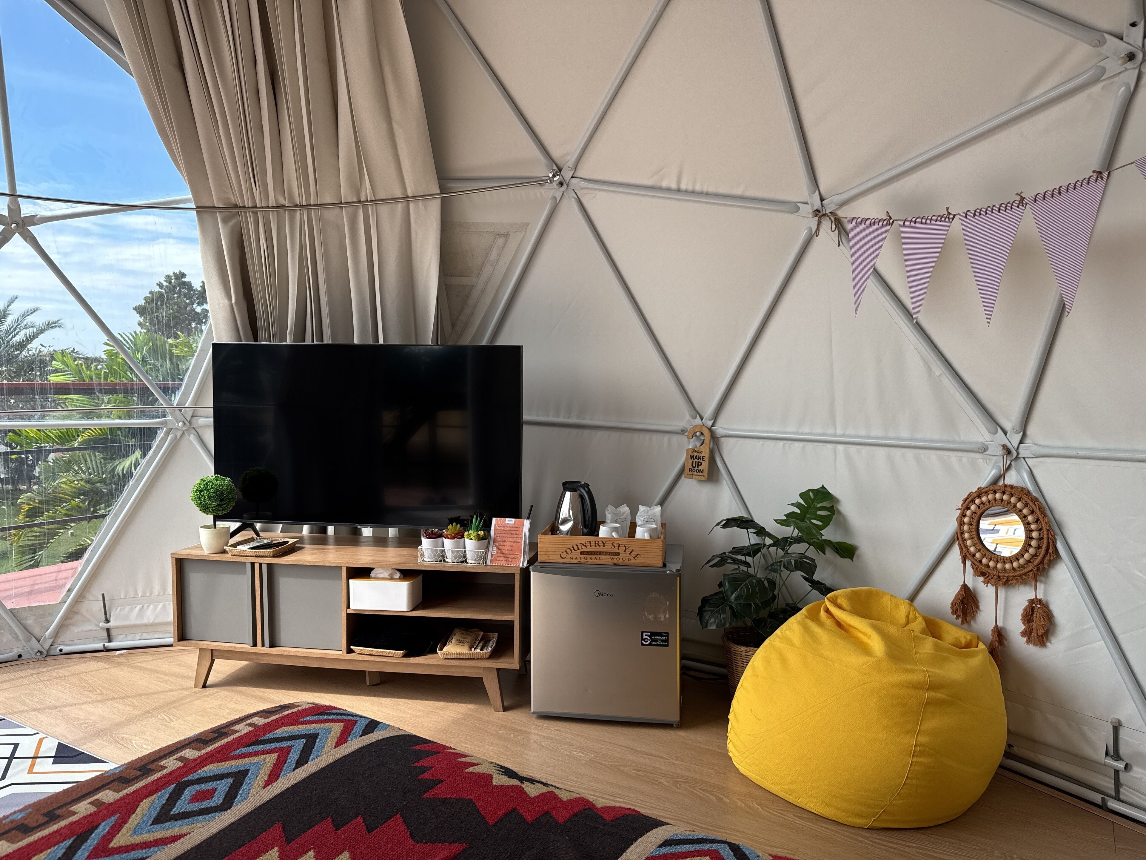 Quarto com tenda, televisão, móveis de madeira, um puff amarelo e vista para o verde lá fora.