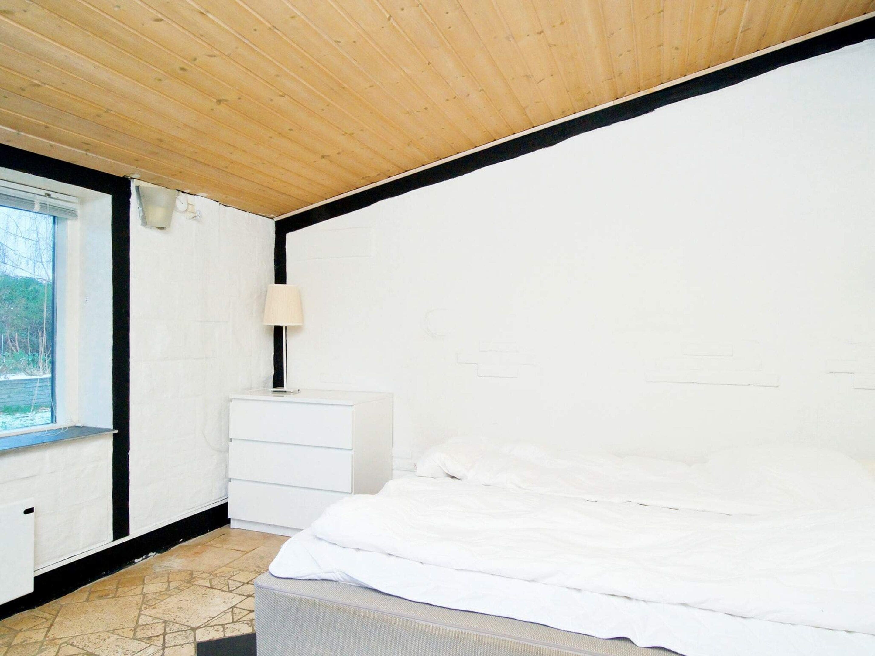 Quarto com teto de madeira, cama branca, mesa de cabeceira com abajur e janela com vista para vegetação.