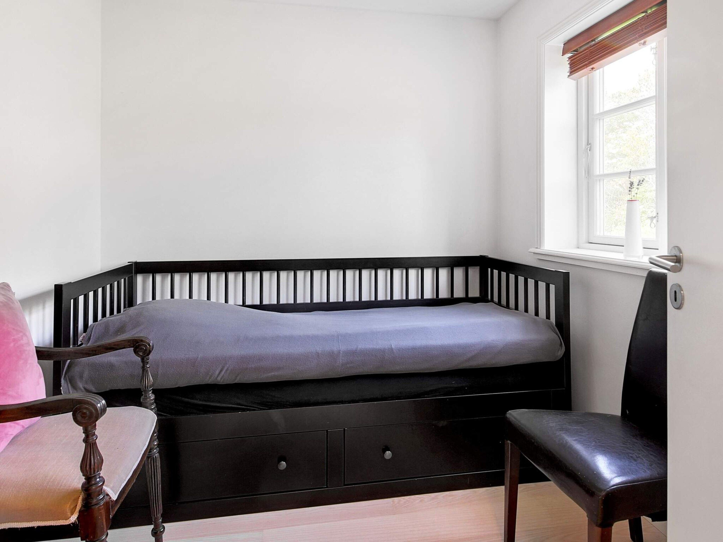 Quarto pequeno e escuro, com uma cama de solteiro, janela, cadeira e mesa de cabeceira.