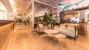 Um lobby moderno com piso de mármore, área de estar com sofás e mesas redondas, e uma parede decorativa com arte abstrata.