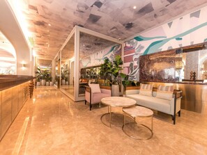 Um lobby moderno com piso de mármore, área de estar com sofás e mesas redondas, e uma parede decorativa com arte abstrata.