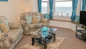 Sala de estar com sofá, mesa de centro de vidro e vista para o mar através de grandes janelas.