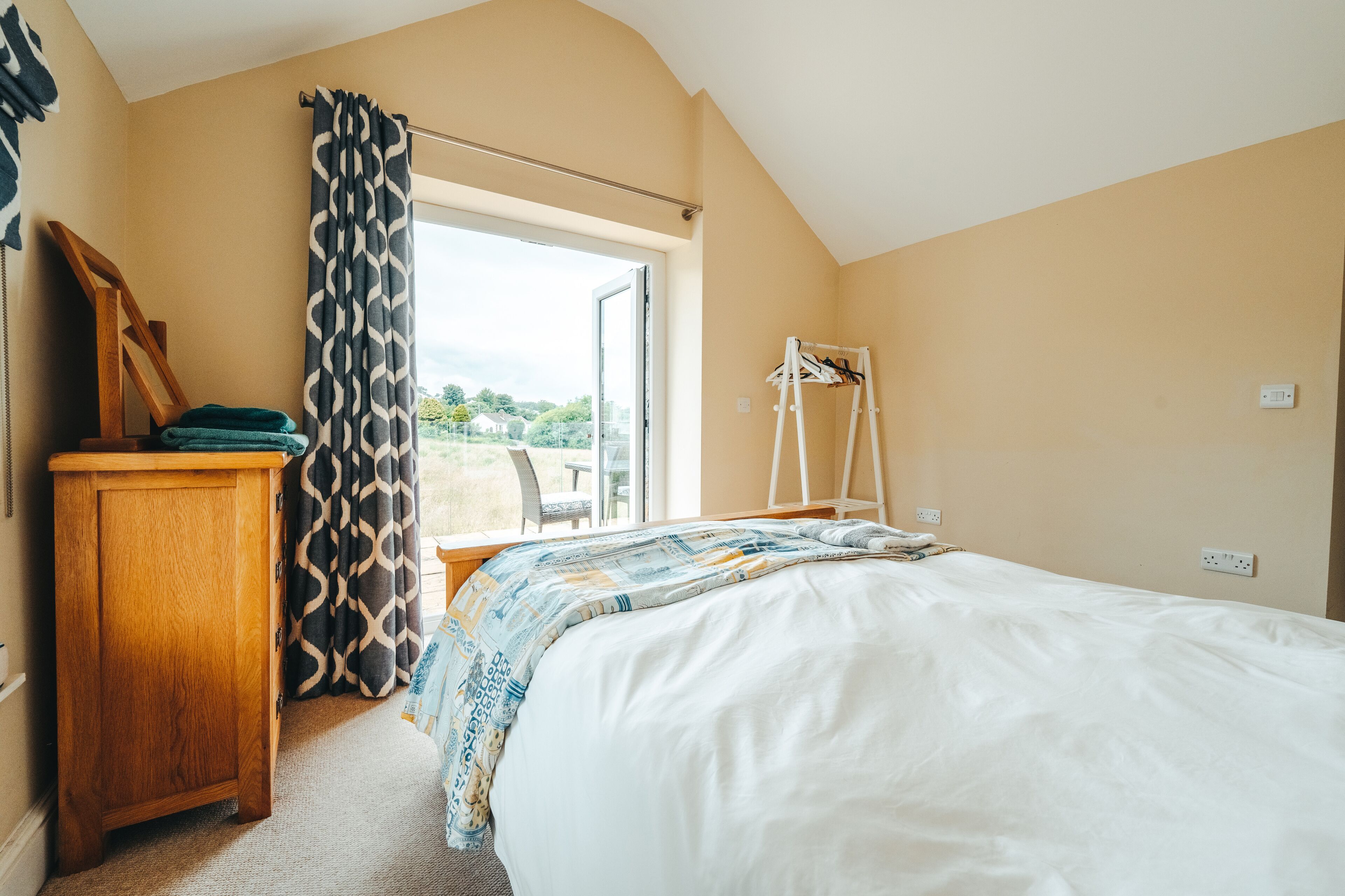 Quarto com cama, um armário de madeira, janela com cortinas e vista para o verde lá fora.