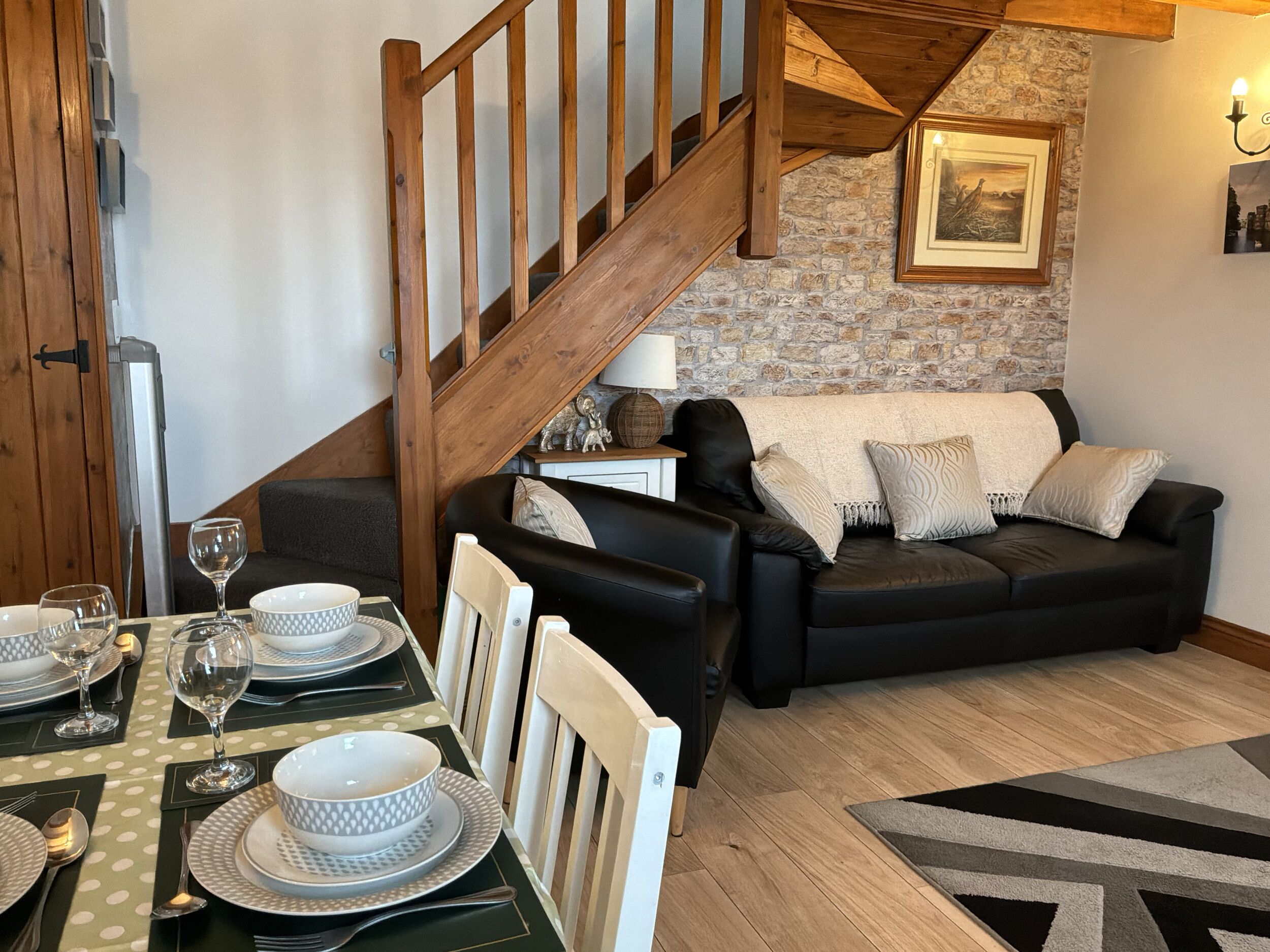 Uma sala de jantar com escada de madeira, parede de pedra, sofá de couro preto e mesa posta com pratos e copos.
