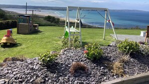 Um parquinho com balanços e estrutura de escalada, um jardim com pedras e plantas, e vista para o mar e colinas ao fundo.