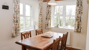 Uma sala de jantar com mesa e cadeiras de madeira, cortinas florais e vista para árvores.