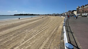 Uma praia com um calçadão pavimentado, um píer ao fundo e edifícios ao longo da costa.
