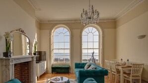 Uma sala de estar ampla com lareira, um sofá verde-água, uma mesa de jantar com cadeiras e grandes janelas com vista para o oceano.