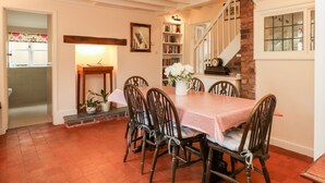 Uma sala de jantar com uma mesa posta para quatro pessoas, rodeada por cadeiras de madeira. Uma estante cheia de livros está à esquerda, e uma escada com uma parede de tijolos à direita.