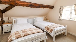 Quarto com duas camas, teto com vigas de madeira, janela com cortinas e mesa de cabeceira com abajur.