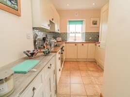Uma cozinha com armários brancos, painel de azulejos, janela com cortina verde e bancada com diversos objetos.