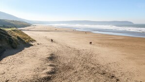 Uma extensa praia de areia com ondas suaves, céu claro e colinas ao longe.