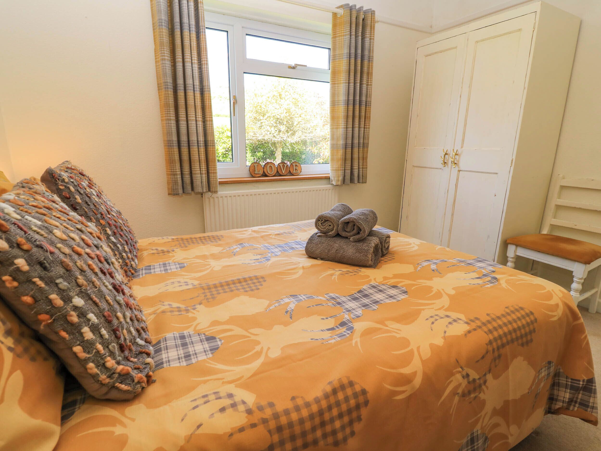 Quarto com cama, janela com cortinas, radiador, guarda-roupa e um banco.