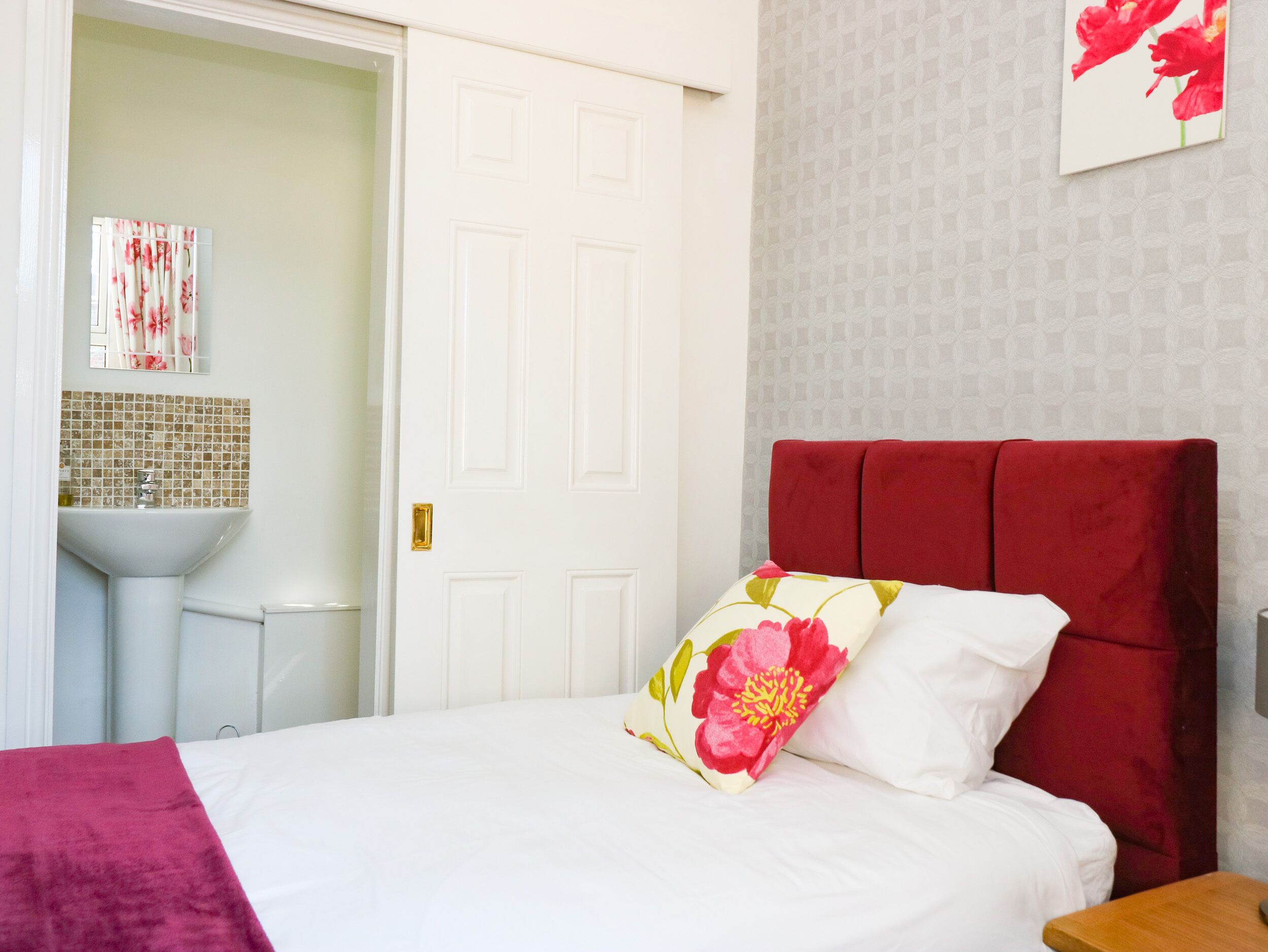 Quarto com cama branca, cabeceira vermelha e almofada floral. Há uma porta branca e uma pia com painel de azulejos.