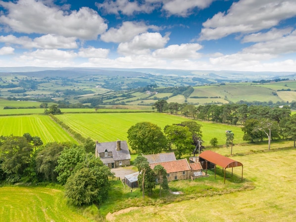 Paisagem rural com campos verdes ondulantes, uma casa de pedra e uma construção com telhado vermelho.