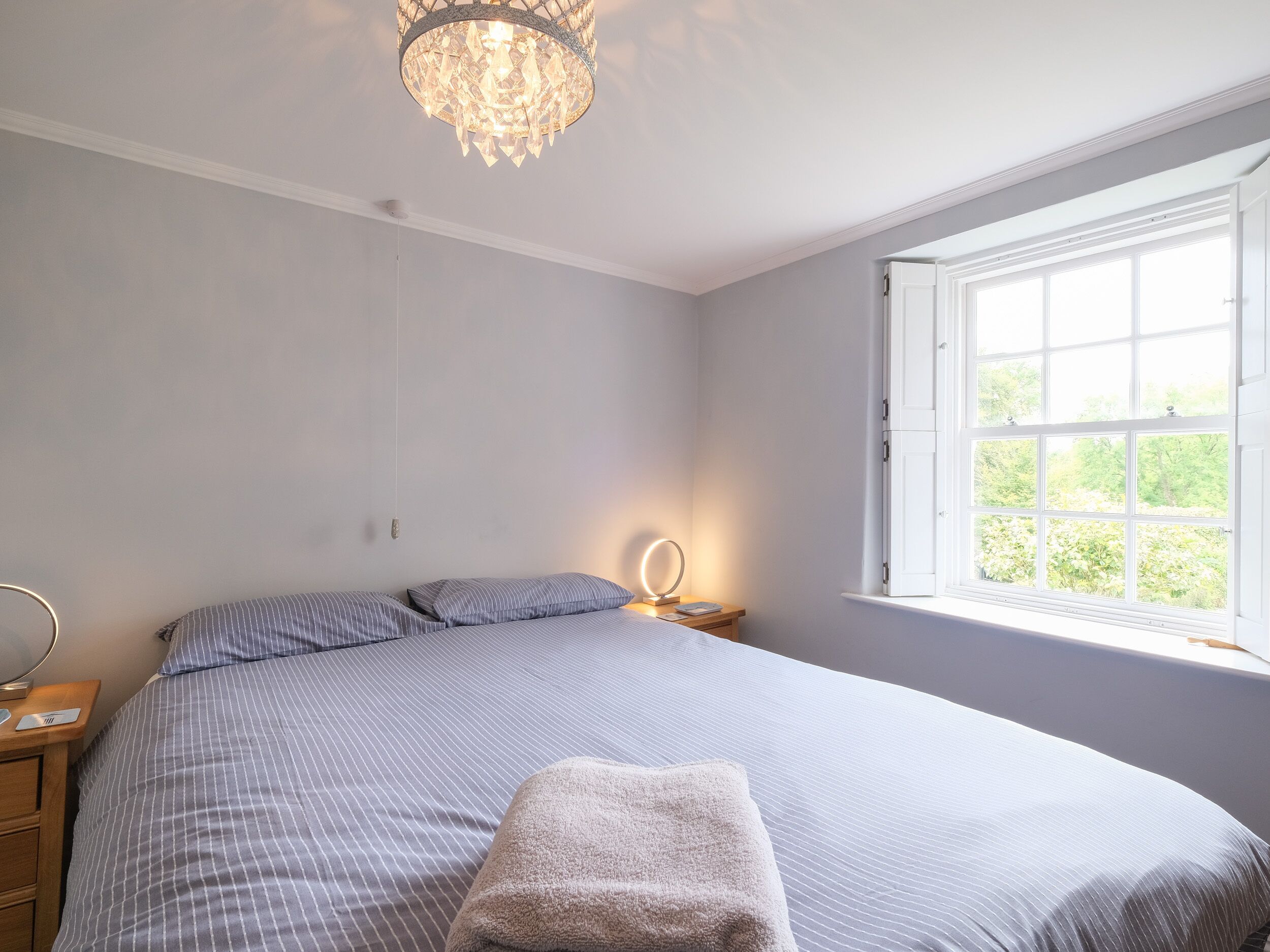 Quarto com cama grande, mesa de cabeceira com abajur, janela com vista para o verde e um lustre.
