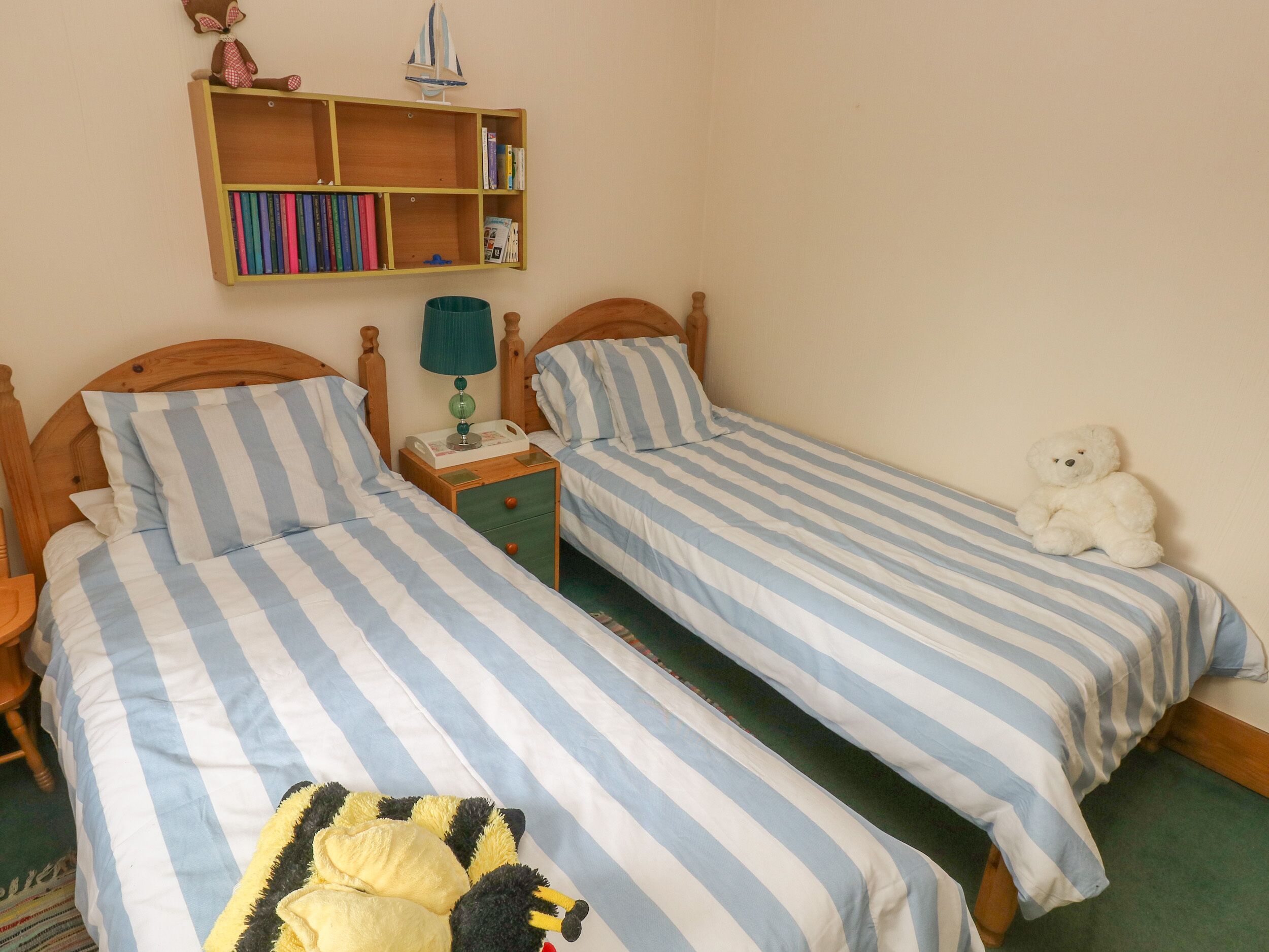 Quarto com duas camas de solteiro, mesa de cabeceira com abajur, estante de livros e um ursinho de pelúcia em uma das camas.