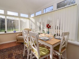 Uma sala de jantar com mesa de madeira posta para quatro, cadeiras brancas com almofadas verdes, um vaso com flores vermelhas e vista para o exterior por meio de persianas brancas.
