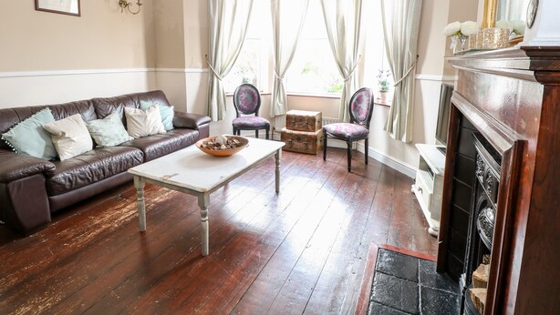 Uma sala de estar com um sofá de couro marrom, uma mesa branca e uma lareira. Há duas cadeiras, uma com padrão floral e outra com estrutura escura. O ambiente possui piso de madeira e grandes janelas com cortinas.