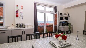 Uma área de jantar com uma mesa posta para dois, um extintor de incêndio e vista para o mar através de uma janela.