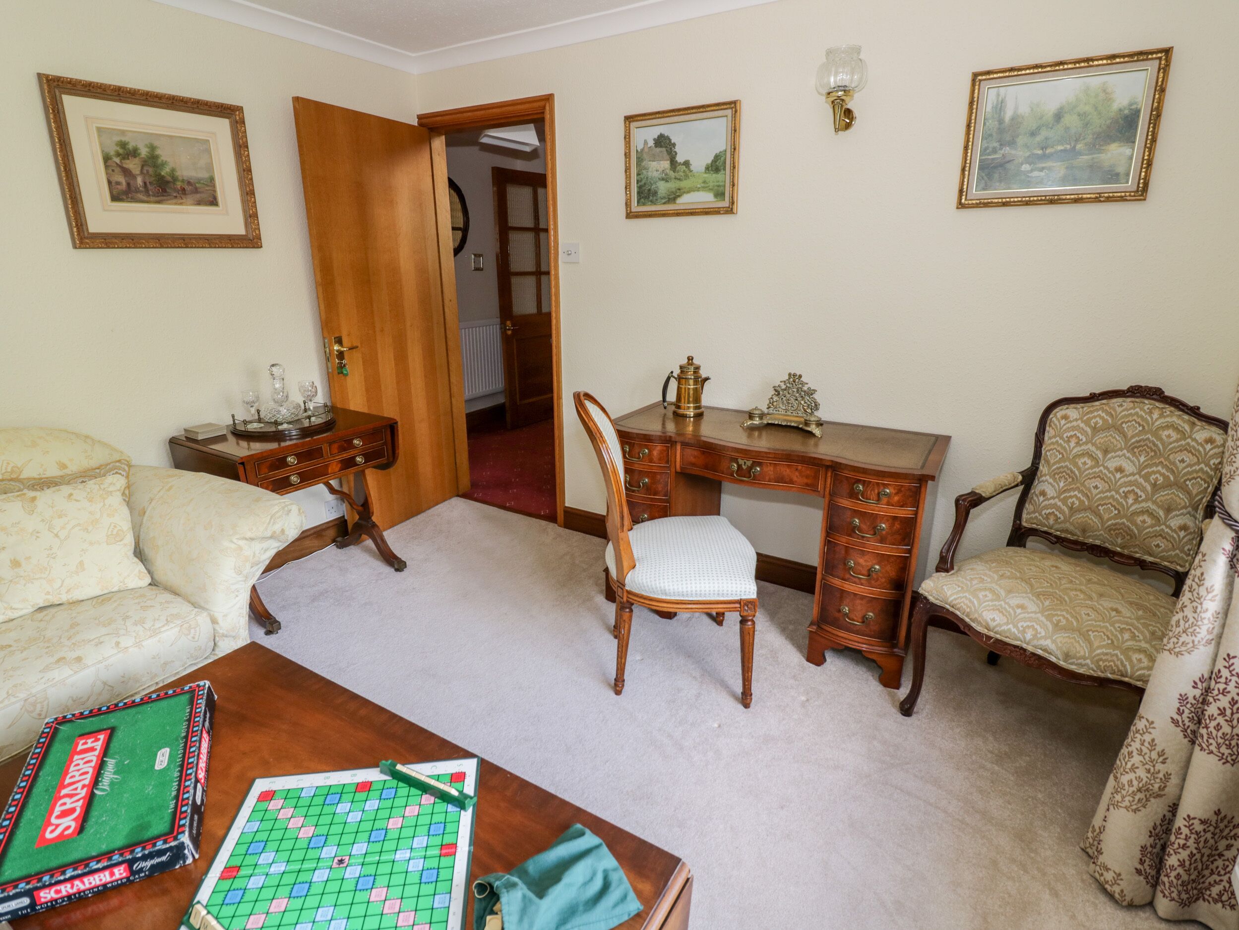 Quarto com escrivaninha de madeira, duas cadeiras, um sofá, mesa de centro com jogo de Scrabble e quadros nas paredes.
