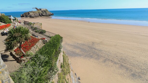 Uma praia costeira com um afloramento rochoso, um calçadão pavimentado e flores vermelhas.
