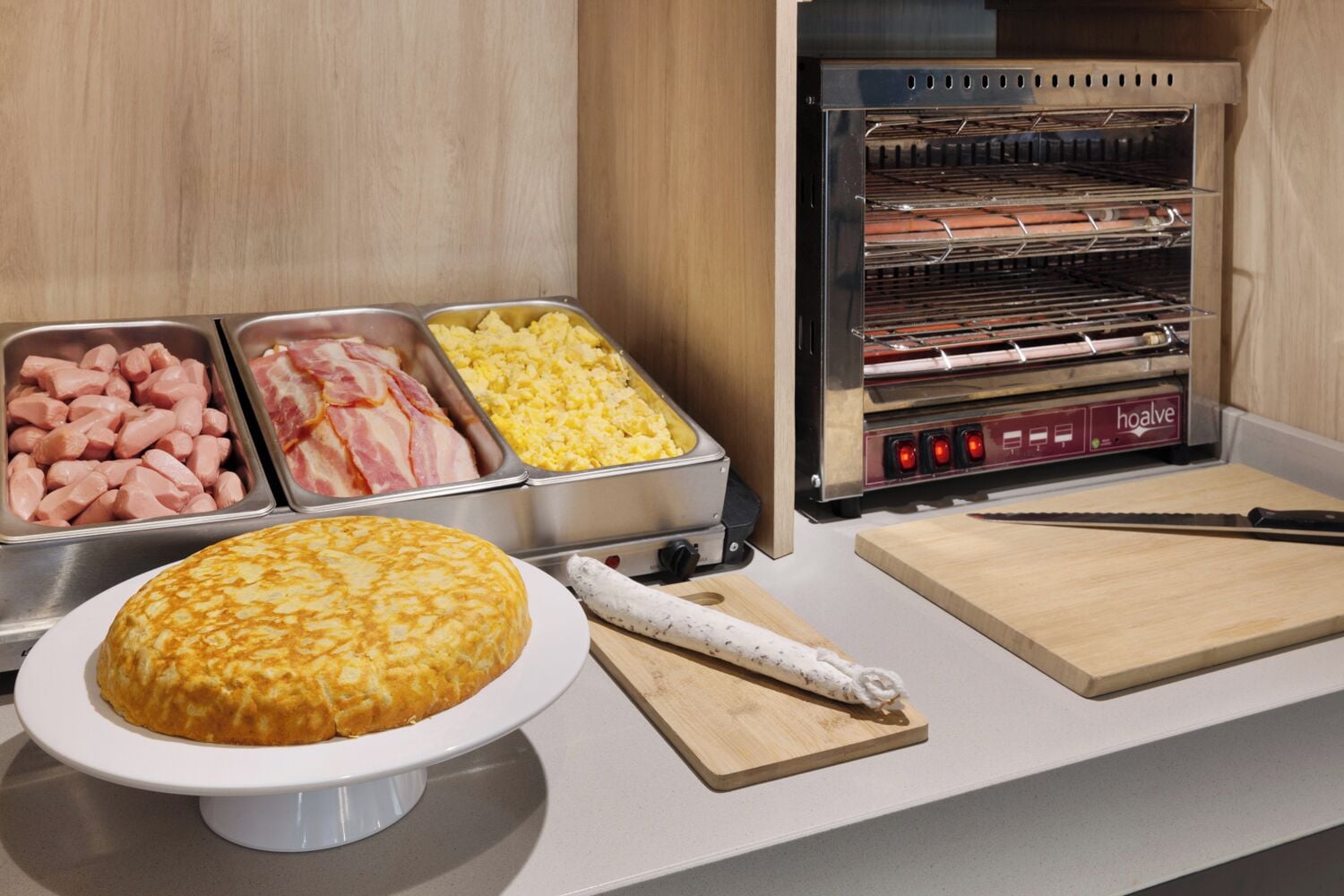 Um buffet de café da manhã com diversas opções de comida, um forno elétrico para torrar pão e uma tábua de corte.