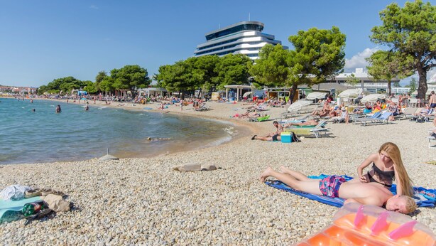 Uma praia de seixos com pessoas tomando sol e nadando, um edifício ao fundo e árvores oferecendo sombra.