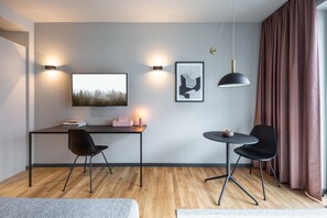 Quarto moderno com escrivaninha, duas cadeiras, mesa redonda, TV na parede, pintura abstrata emoldurada e luminária pendente.