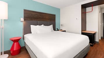 Quarto de hotel moderno com uma cama grande, uma mesa lateral vermelha de destaque, um abajur e uma mesinha de cabeceira de madeira.