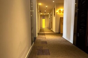 Corredor de hotel com piso revestido de carpete, iluminação embutida e uma porta à direita.
