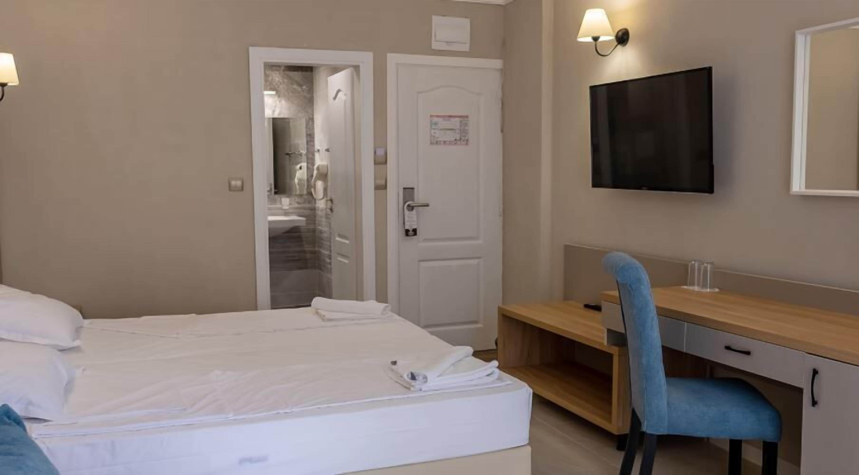Quarto de hotel com uma cama, uma escrivaninha, uma cadeira, uma televisão e uma porta.