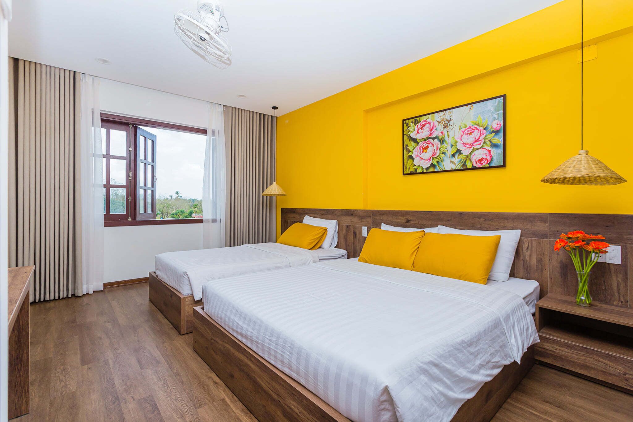 Quarto de hotel com duas camas, uma janela grande com cortinas, uma cabeceira de madeira e um quadro de flores na parede.