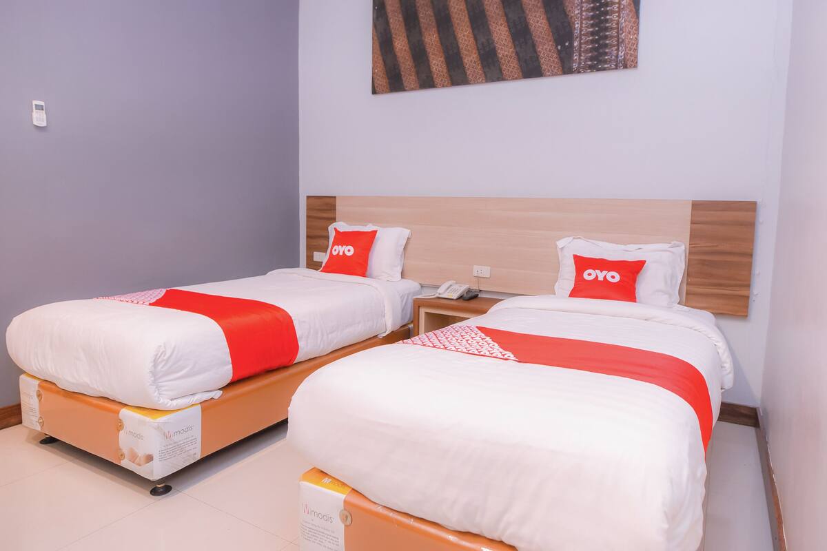 Duas camas com roupa de cama branca e vermelha, uma cabeceira de madeira, uma mesinha de cabeceira e um telefone fixado na parede.