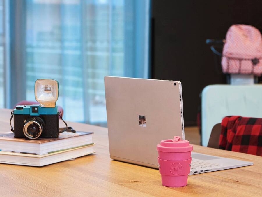 Um laptop, um copo rosa e uma câmera antiga sobre uma mesa de madeira.