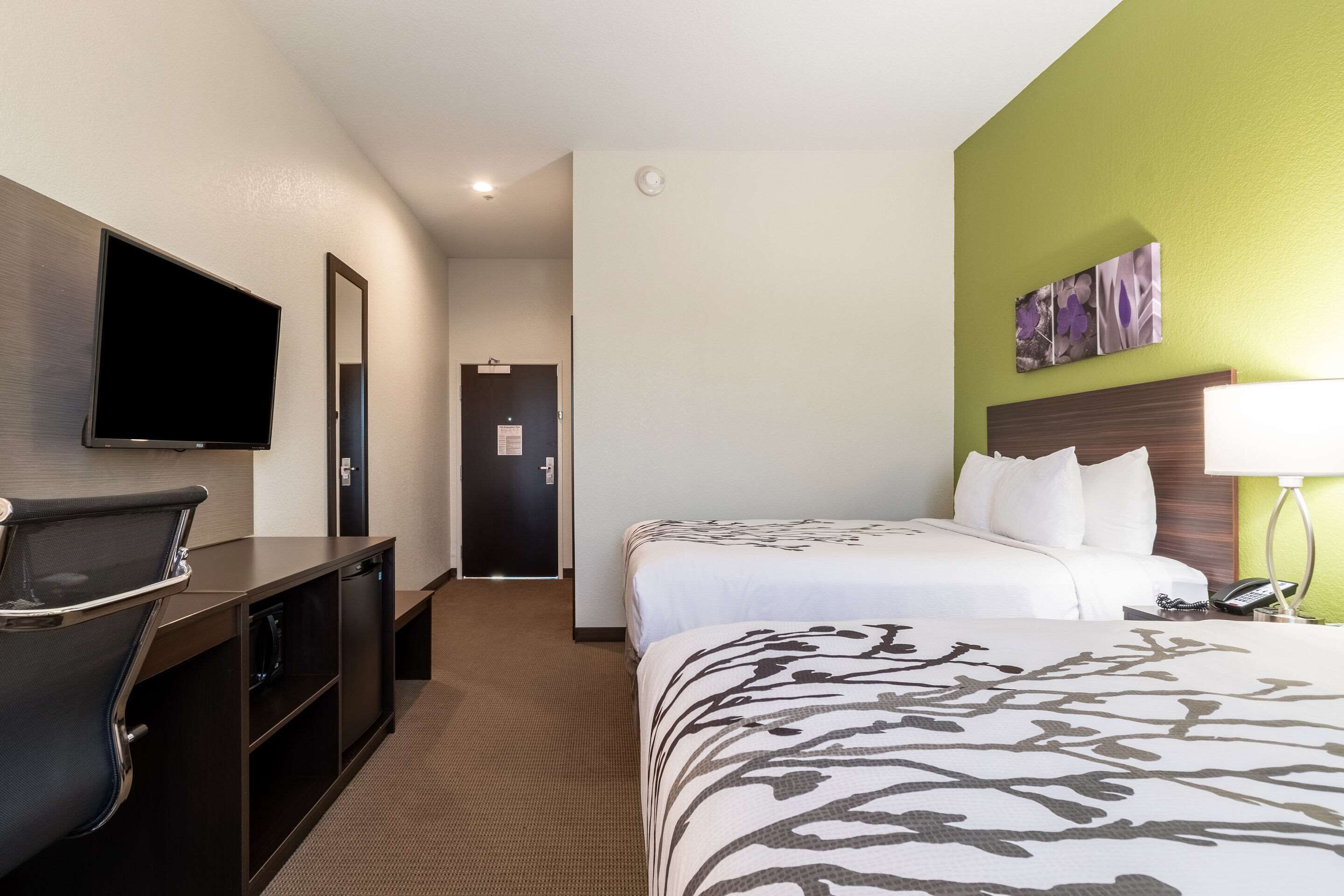 Quarto de hotel com cama, escrivaninha com cadeira, televisão e um quadro na parede.