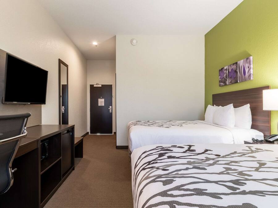Quarto de hotel com cama, escrivaninha com cadeira, televisão e um quadro na parede.
