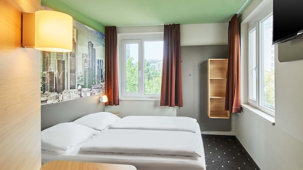 Quarto de hotel com duas camas, uma escrivaninha, televisão, janela com cortinas e um mural com vista para a cidade.