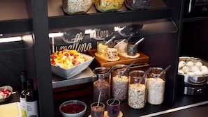 Uma área de buffet com diversos alimentos, incluindo uma tigela de salada de frutas, potes com nozes e sementes, e uma tigela de compota de frutas vermelhas.