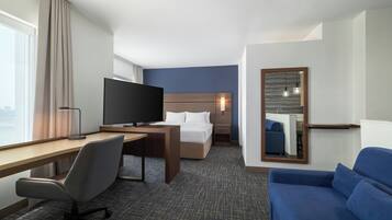 Quarto de hotel com uma cama, uma escrivaninha com televisão, uma cadeira, um sofá azul e um espelho.