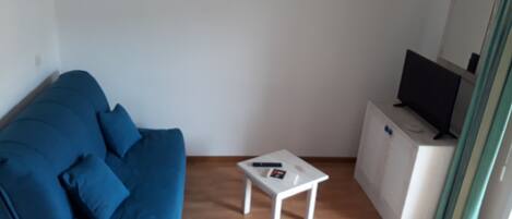Quarto com sofá azul, mesa pequena branca, televisão num móvel e janela com cortinas.